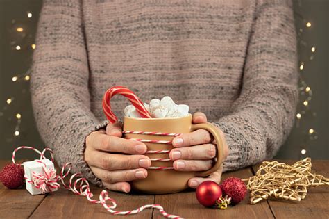 나무 테이블에서 따뜻한 양모 스웨터를 입은 인식 할 수없는 여성은 뜨거운 음료 마시멜로 및 사탕 지팡이와 함께 갈색 아늑한 머그잔을 들고 있습니다 크리스마스 조명 켜짐 선물