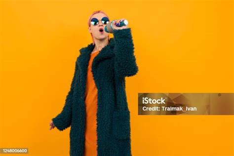 어두운 코트와 선글라스에 짧은 분홍색 머리를 가진 특별한 아름다운 여자는 오렌지 배경에 고립 된 마이크에 노래 갈색 머리에 대한 스톡 사진 및 기타 이미지 Istock