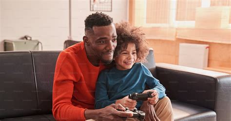 가족 여가 시간 행복한 다인종 면도하지 않은 아버지와 그의 곱슬곱슬한 세련된 아들은 조이스틱으로 콘솔에서 비디오 게임을 하면서 좋은 감정으로 집에서 주말을 즐깁니다 스톡