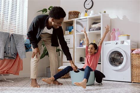 세탁실 욕실에서 집안일을하는 동안 아버지와 함께 놀아요 한 남자가 세탁기에서 옷을 풀고 나서 딸의 그릇을 잡아 당겨 재미있게 놀 수 있습니다 빨래에 대한 스톡 사진 및 기타