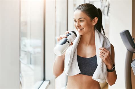 휴식 시간 체육관에서 창 앞에 서 있는 동안 그녀의 어깨에 물을 마시는 흰색 수건을 가진 스포츠웨어에서 젊고 쾌활 한 여자