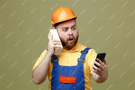 Запутавшись, держа телефон, кладя кирпич на голову, молодой строитель ...