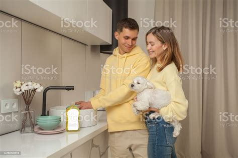 부엌에서 쾌활한 커플의 초상화 싱크대에서 설거지를 하는 남자 손에 작은 흰색 개를 들고 있는 여자 테이블에 빈 라벨이 있는 식기 세척액 명랑한에 대한 스톡 사진 및 기타