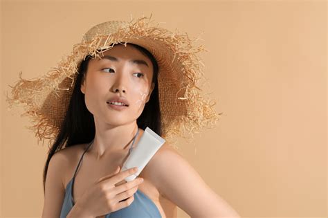 얼굴에 자외선 차단제를 바르고 있는 밀짚모자를 쓴 아름다운 젊은 여성이 베이지색 배경에 자외선 차단 크림을 들고 있다 햇볕 차단 로션에 대한 스톡 사진 및 기타 이미지