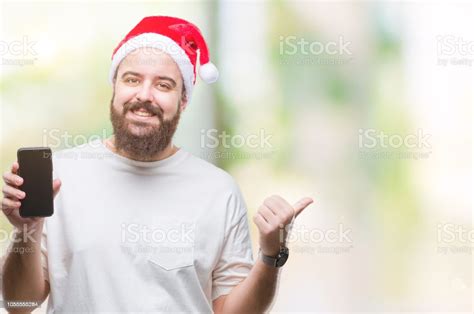 젊은 백인 남자 고 행복 얼굴 미소와 측면까지 손가락으로 보여주는 격리 된 배경 위에 크리스마스 모자 보여주는 스마트폰 화면을 입고 가리키기에 대한 스톡 사진 및 기타