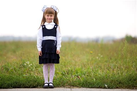 흐리게 밝은 녹색 햇볕이 잘 드는 잔디와 흰색 하늘 복사 공간 배경에 긴 금발 머리에 학교 유니폼과 흰색 활귀여운 사랑스러운 심각한 사려 깊은 1 학년 소녀의 전체 길이