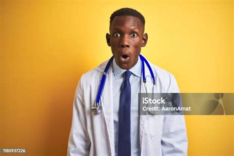 고립 된 노란색 배경 위에 서있는 청진기를 입은 아프리카 계 미국인 의사가 놀라움의 표정 두려움 및 흥분된 얼굴로 충격을 받았습니다 의사에 대한 스톡 사진 및 기타 이미지