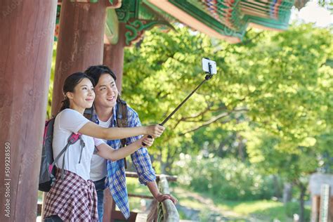 한국의 옛날 전통 가옥의 처마와 녹색 숲을 배경으로 여행하며 스마트폰으로 다양한 포즈를 셀카로 찍는 젊은 아시아 한국 남녀 모델 фотография Stock Adobe