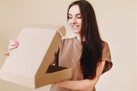 미소 짓는 갈색 머리 여성이 베이지 색 배경에 갈색 티셔츠를 입은 상자를 들여다 봅니다 감정에 대한 스톡 사진 및 기타 이미지 감정 건강에 좋지 않은 음식 건강하지