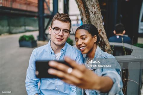 쾌활한 아프리카계 미국인 여성이 도시 환경에 앉아 인터넷 블로그를 위한 사진을 만드는 백인 힙스터 남자와 함께 스마트폰 전면 카메라에 포즈를 취하고 있으며 셀카를 찍는