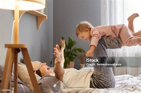 매우 행복한 기뻐하는 어머니의 초상화는 아기 딸과 함께 시간을 보내고 침대에 누워 침실에서 놀고 엄마의 다리로 날아가는 아이와 웃고 있습니다 18 23 개월에 대한 스톡