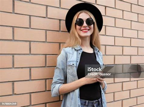 검은 둥근 모자를 쓰고 카메라를 보고 있는 전화로 미소짓는 젊은 여성의 초상화 벽돌 벽 배경 위에 청바지 재킷 20 29세에 대한 스톡 사진 및 기타 이미지 Istock