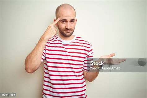 흰색 고립 된 배경에 캐주얼 스트라이프 빨간 티셔츠를 입고 수염을 가진 젊은 대머리 남자는 혼란과 복사 공간을 보여주는 열린 손바닥과 짜증이 이마에 손가락을 가리키는 그것에