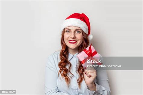 곱슬 머리와 범본과 빨간 모자를 가진 산타 클로스 소녀는 빨간색 새틴 리본 활과 선물 상자를 보유하고 회색 벽의 배경에 미소 사이트에 대한 메리 크리스마스와 새해 웹 배너