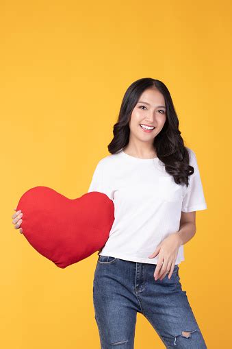 행복한 아시아 젊은 여성은 노란색 배경에 고립 된 귀엽고 사랑스러운 미소 빨간 마음을 들고 긴 곱슬 머리 사랑과 발렌타인 데이