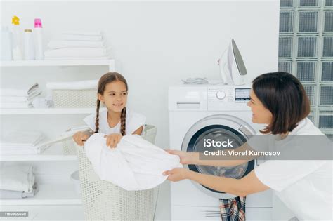 어두운 짧은 머리를 가진 행복한 여자는 바구니에서 빨래를 꺼내 행복한 아이는 그것에 포즈 세탁기 와 상단에 철 접힌 흰색 수건으로 선반 근처 욕실에서 시간을 보내고 세탁