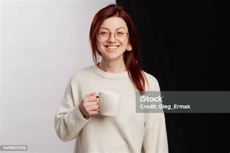 뜨거운 음료가 활기를 띠고 있습니다 차 또는 커피 한 잔을 손에 들고 있는 빨간 머리 여자 큰 안경에 반쯤 흰색과 검은 색 배경에 얼굴 표정이 서 있습니다 개성 개념에 대한