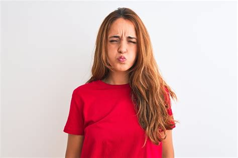 젊은 빨간 머리 여자 빨간 캐주얼 티셔츠 를 입고 흰색 고립 된 배경 퍼핑 뺨을 통해 앉아 재미있는 얼굴 공기 미친 표정으로 입이 팽창 T 셔츠에 대한 스톡 사진 및 기타