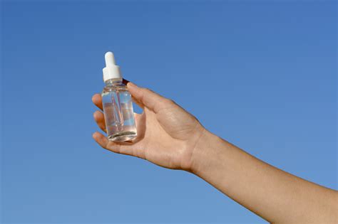 푸른 하늘 배경에 세럼이 든 투명한 유리 항아리를 들고 있는 여성의 손 미용 제품의 개념 주름을 매끄럽게 하고 얼굴에 수분을 공급 영양 및 관리 25 29세에 대한 스톡