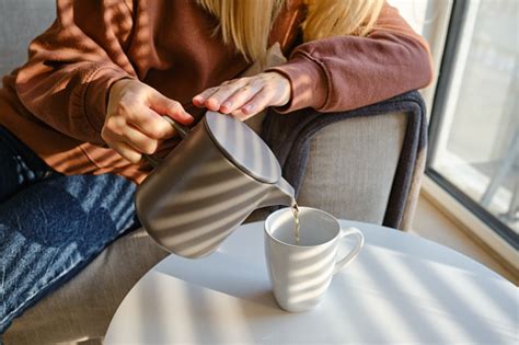 소녀는 찻주전자에서 컵에 뜨거운 차를 붓습니다 가정의 안락함을 즐기십시오 창문에서 태양광선에 있는 한 여성의 손에 하얀 컵을 클로즈업합니다 여자의 손에 차 한 잔 겨울에