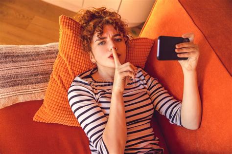 Ginger Cute Girl Lying On The Sofa With A Phone In Hand And Showing A Silence Sign Stock Image