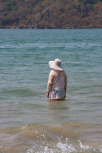 알아볼 수 없는 백인 여성의 이미지는 바닷물 속에 허벅지 깊숙이 서 있고 태양 보호복을 입고 플로피 태양 모자와 태양 덮개를 씌우고 뒷모습을 하고 있다 50 54세에 대한