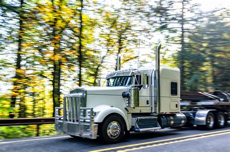 흐릿한 나무가 있는 좁은 가을 도로를 달리는 플랫 베드 세미 트레일러에 고정된화물을 운반하는 그릴 가드가 있는 클래식 빅 리그 화이트 세미 트럭 트랙터 0명에 대한 스톡