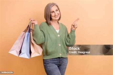 카키색 가디건을 입은 쾌활한 노인 여성의 사진은 베이지색 배경에 고립된 빈 공간을 나타내는 새 옷을 들고 있다 가리키기에 대한 스톡 사진 및 기타 이미지 Istock