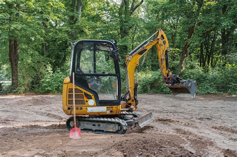 빨간 삽이 기대어 있는 작은 노란색 크롤러 굴착기가 푸른 숲의 나무로 둘러싸인 모래 공사 현장에 주차되어 있습니다 0명에 대한