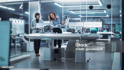 하이테크 시설에서 일하는 두 명의 여성 로봇 공학 엔지니어와 기계 운영자는 기술 스타트업 실험실에서 랩톱 컴퓨터를 사용하여 로봇