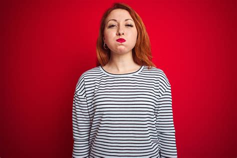 젊은 빨간 머리 여자 를 입고 스트랩 해군 셔츠 위에 서 빨간 고립 된 배경 퍼핑 뺨 재미 있는 얼굴 공기 미친 표정으로 입이 팽창 T 셔츠에 대한 스톡 사진 및 기타