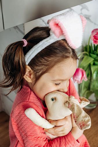 분홍색 드레스와 토끼 귀를 입은 작은 검은 머리 소녀가 집에서 부엌 의 집 꼭대기에 앉아있는 장난감 토끼를 껴안습니다 행복한 부활절 개념 4 5세에 대한 스톡 사진 및 기타