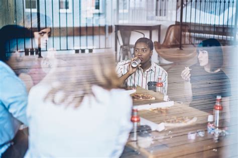 캐주얼 한 옷을 입은 다문화 젊은 이들이 술을 마시고 현대 아파트의 유리 벽 뒤에 테이블에 앉아있는 파티에서 피자를 먹습니다 다양한 힙스터 남자들이 함께 쉬고 있습니다