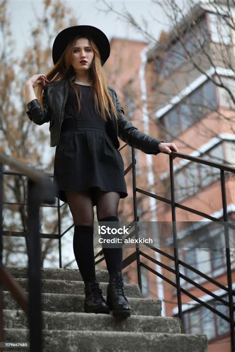 한 소녀가 전화기를 들고 가죽 조끼를 입고 도시 거리를 걷고 있다 모자와 어두운 메이크업 외부젊은 아름다운 소녀 거리에서 고딕