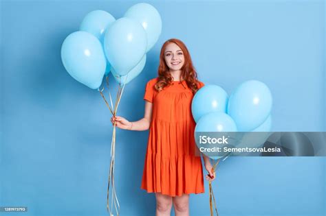 풍선 우아한 드레스를 입고 빨간 물결 모양의 머리를 가진 예쁜 여자는 카메라를 보고 그녀의 생일을 축하하는 행복한 여자 파란색