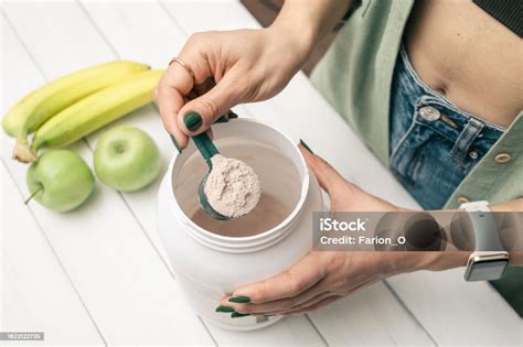 청바지와 셔츠를 입은 여자가 셰이커 바나나 사과 과일이 있는 흰색 나무 테이블 위의 플라스틱 항아리 위에 유청 단백질 가루 부분이 담긴 계량 스푼을 들고 있습니다 단백질