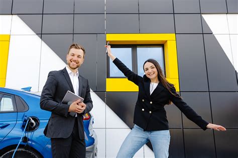 행복한 여성 고객이 새 차에 서서 야외 대리점의 딜러로부터 열쇠를 가져갑니다 여자는 열쇠를 손에 들고 새 차에 매우 만족합니다