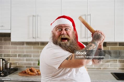 산타클로스 모자를 쓰고 문신을 한 남자가 흰색 티셔츠를 입고 야구 방망이처럼 나무 롤링 핀을 손에 들고 카메라를 바라보며 기쁨 만족도 불운 놀라움 장난기 등 그의 감정을