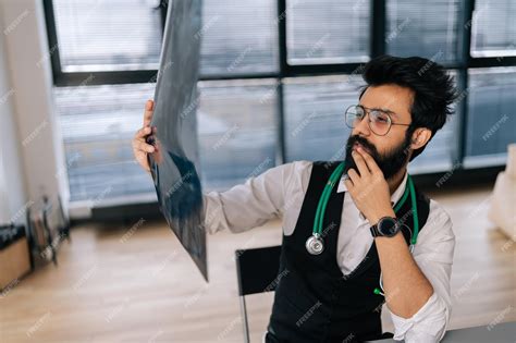 Mri 이미지 스캔을 보고 진단을 생각하는 안경을 쓴 사려 깊은 인도 남성 의사 병원에서 척추 엑스레이 이미지를 검사하는 방사선과 의사 프리미엄 사진