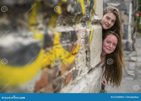 Two Teen Girls Look Out From Behind The Corner Of A Stone House Stock