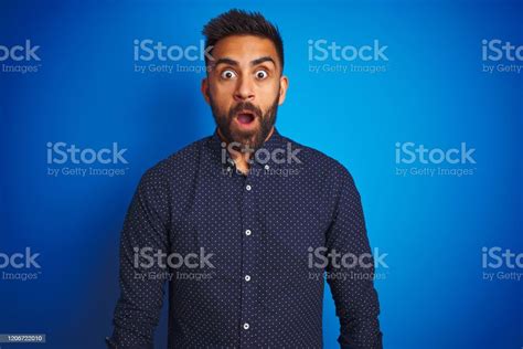 고립 된 파란색 배경 위에 서있는 셔츠를 입은 젊은 인도의 우아한 남자는 놀라움의 표정 두려움과 흥분된 얼굴로 충격을 받았습니다 경영자에 대한 스톡 사진 및 기타 이미지