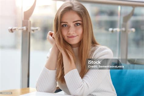 즐거운 표정으로 쾌적 한 모습의 여자의 헤드샷 캐주얼 한 옷을 입은 카메라를 바라보 고 카페테리아에서 편한 소파에 앉아서 질서를 기다리며 공공 장소에서 자유 시간을 보낸다