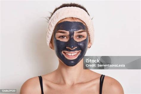 얼굴에 검은 점토 얼굴 마스크를 가진 아름다운 웃는 여자 행복한 표정으로 카메라를보고 그녀의 머리에 헤어 밴드를 가지고 흰색 배경에 고립 된 포즈 18 19세에 대한 스톡