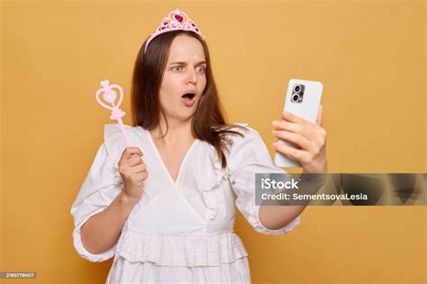 하얀 공주 의상과 티아라를 착용한 여성이 스마트폰을 보며 놀라움을 표현하며 밝은 노란색 배경에 장난기 넘치는 순간을 보여줍니다 30 34세에 대한 스톡 사진 및 기타 이미지