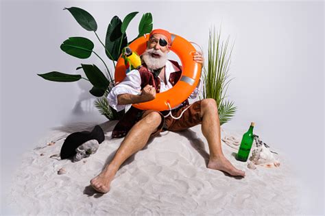 생명 반지와 화려한 앵무새와 함께 모래 섬에 앉아있는 기발한 해적 할아버지는 해적 의상을 입고 유머와 모험을 발산합니다 10월에 대한 스톡 사진 및 기타 이미지 Istock