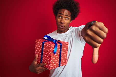 화가 얼굴 엄지 손가락으로 싫어하는 것을 보여주는 부정적인 기호 거부 개념으로 고립 된 빨간 배경 위에 서있는 생일 선물을 들고 있는 젊은 아프리카 계 미국인 남자 선물에
