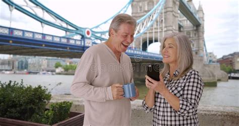 Old Caucasian Wife Takes Photo Of Husband On Phone Near Tower Bridge In