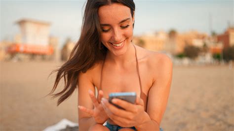 긴 머리카락을 가진 슬림 한 아름다운 갈색 머리 여성은 해변에 앉아있는 휴대 전화를 사용하고 있습니다 스마트 폰으로 정보를 스크롤하는 화려한 웃는 소녀 여자에 대한 스톡