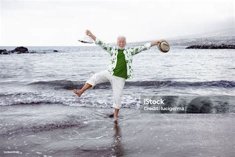 해안선에 맨발로 서서 농담으로 한쪽 다리를 들어 올리는 노인의 행복 해변을 즐기는 손에 신발과 모자를 들고있는 성숙한 남자 편안하고 즐거운 은퇴자의 개념 70 79세에 대한