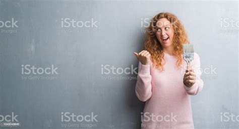 페인트 브러시 고 보여주는 행복 한 얼굴은 웃 고 옆까지 엄지를 들고 회색 그런 지 벽에 빨강 머리 젊은 여자 가리키기에 대한 스톡 사진 및 기타 이미지 Istock